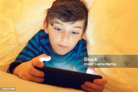 행복 웃는 소년은 담요 아래 침대에 앉아 어둠 속에서 게임에서 스마트 폰에서 재생 합니다 아이의 얼굴은 밝은 모니터로 비춰 집니다 소년에 대한 스톡 사진 및 기타 이미지