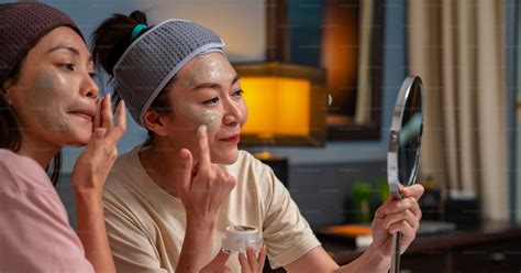 웃는 아시아 여자 친구들은 침대에 앉아 집에서 함께 얼굴에 스킨 케어 페이셜 마스크를 바르고 있다 여성 게이 커플은 긴장을 풀고 행복과 함께 뷰티 페이셜 트리트먼트를