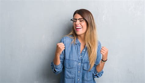 젊은 성인 여자 입고 안경 매우 행복 하 고 흥분 무기와 우승자 제스처를 하 고 그런 지 회색 벽에 웃 고 성공을 위한 비명 축 하 개념입니다 긍정적인 감정 표현에 대한