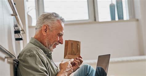 낙관적인 노인이 노트북 화면 앞에서 작은 선물 가방을 들고 적극적으로 몸짓을 하고 있다 사진 Unsplash의 하루 이미지