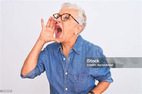데님 셔츠와 안경을 쓰고 고립된 흰색 배경 위에 회색 머리의 선임 여성이 입을 다물로 큰 소리로 외치고 비명을 지르고 있습니다 통신 개념입니다 노인에 대한 스톡 사진 및