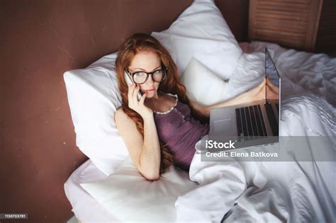 침대에 앉아있는 빨간 머리 소녀 그녀는 전화로 말하고 노트북에서 뭔가를 하 고 있습니다 그녀는 카메라를 보고 있어요 전면 보기