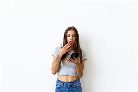 충격을 받은 여성이 휴대전화를 사용하여 흰 벽 배경 위에 고립된 포즈를 취하고 있는 사진 Brand Name Smart Phone에 대한 스톡 사진 및 기타 이미지