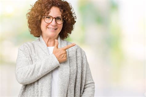 손과 얼굴에 행복 하 고 자연 식 가진 측까지 손가락으로 가리키는 얼굴의 미소와 쾌활 한 고립 된 배경 위에 재킷과 안경을 착용 해 아름 다운 중간 어른 고위 여자