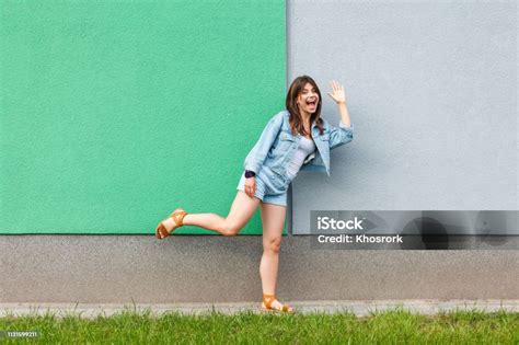 캐주얼 청바지 데님 스타일의 행복 한 흥분 아름 다운 여자의 전체 길이 초상화 여름과 밝은 파란색 벽 근처에 서 서 카메라 인사말을 보고 그녀의 손을 흔들며 분리에 대한