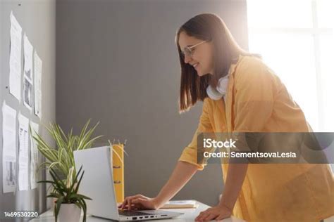 노트북 컴퓨터를 사용하고 사무실의 테이블 근처에 서서 일을 즐기고 노트북을 마무리하고 끄는 노란색 셔츠를 입은 바쁜 웃는 여성의