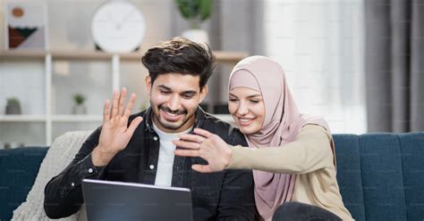 웃고 있는 쾌활한 젊은 무슬림 커플은 집에서 노트북을 들고 소파에 앉아 친척이나 친구와 화상 통화를 하면서 손을 흔든다 가족 기술 와이파이 인터넷 개념 사진