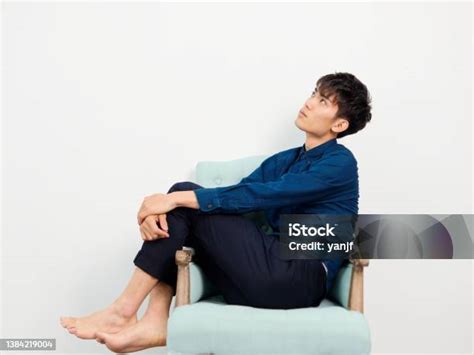 안락의자에 앉아 흰색 벽 배경에 포즈를 취하는 파란색 셔츠에 곱슬 검은 머리를 가진 잘 생긴 중국 청년의 초상화 무릎에 손을 교차 전체 길이 측면 보기 스튜디오 샷 남자에