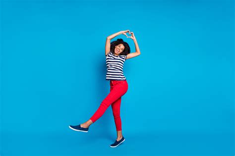 전체 길이 바디 사이즈 사진 의 쾌활 한 긍정적인 좋은 예쁜 귀여운 여자 친구 착용 바지 바지 스트라이프 티셔츠 손 을 보여주는 심장 모양 기호 스테핑 고립 된 밝은 파란색