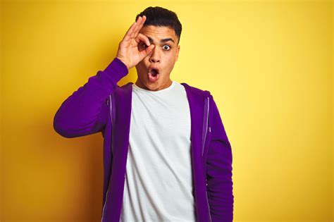 고립 된 노란색 배경 위에 서있는 보라색 스웨트 셔츠를 입은 젊은 브라질 남자는 손가락을 통해 보고 있는 놀란 얼굴로 충격을 받은 확인 제스처를 하고 있습니다 믿을 수 없는