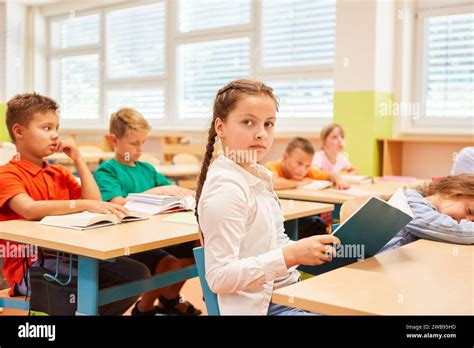 Side View Portrait Of Schoolgirl Holding Book While Sitting On Bench With Friends In Classroom