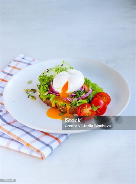 통밀 빵에 묽은 수란을 얹고 샐러드 토마토 적양파 양상추 오이를 곁들인 요리 방울토마토 마이크로그린 참깨 호박씨를 면 체스판 패턴 냅킨에 흰색 접시에 장식합니다 건강한