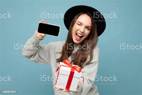 매력적인 행복한 즐거운 젊은 갈색 머리 여자 는 유행 검은 모자와 캐주얼 회색 스웨터를 입고 파란색 배경 벽에 고립 된 모형을위한 휴대 전화 화면 디스플레이를 보여주는 선물