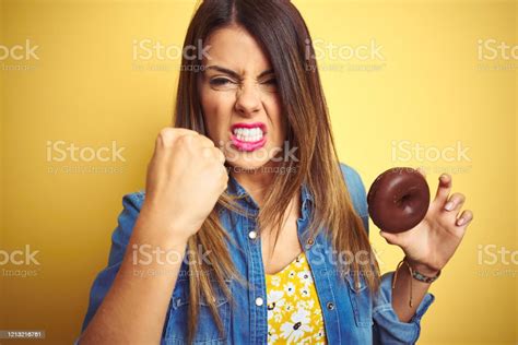 노란색 배경에 초콜릿 도넛을 먹는 젊은 아름다운 여자는 화가 좌절 분노와 함께 소리 미친 고함 제기 손으로 고함 분노 개념