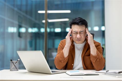 두통 편두통 사무실 안에서 직장에서 머리에 손을 얹고 아픈 남자 노트북을 들고 책상에 앉아 고통받는 아시아 남자 슬픈 회사원 걱정하는에 대한 스톡 사진 및 기타 이미지